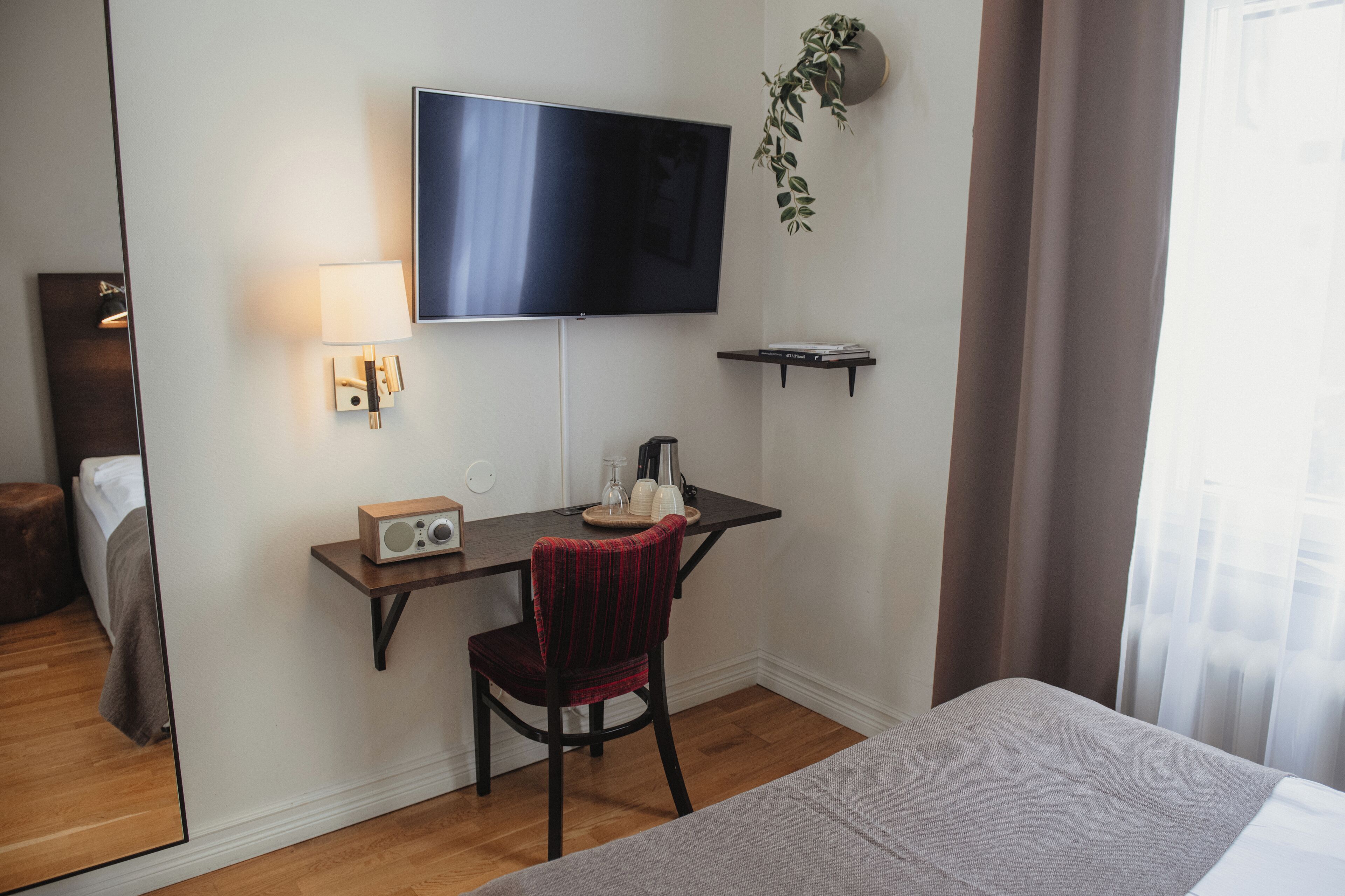 Quarto de hotel com cama, escrivaninha com cadeira, televisão de tela plana, abajur, rádio, cafeteira, prateleira pequena e janela com cortinas.