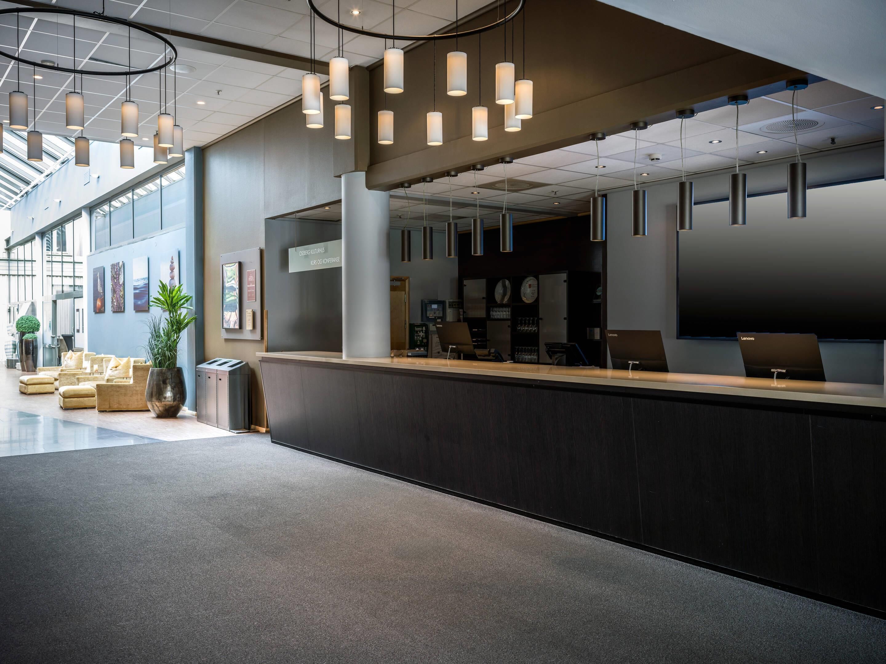 Uma área de recepção moderna com um balcão escuro, diversas luminárias pendentes e um espaço de estar com sofás e uma grande planta em vaso.