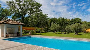 Piscina com deck de madeira, um pequeno gazebo e área gramada com árvores e cerca.