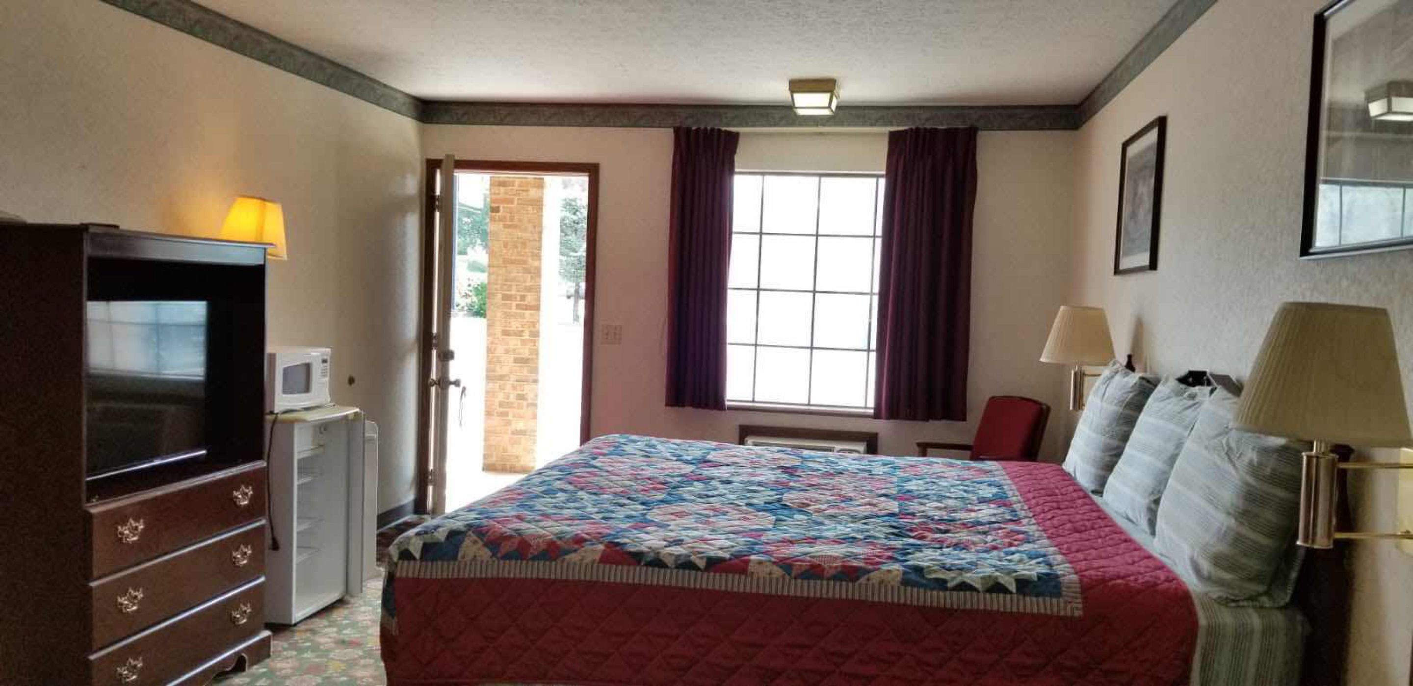 Quarto de hotel com cama, televisão, escrivaninha com luminária e janela com cortinas.