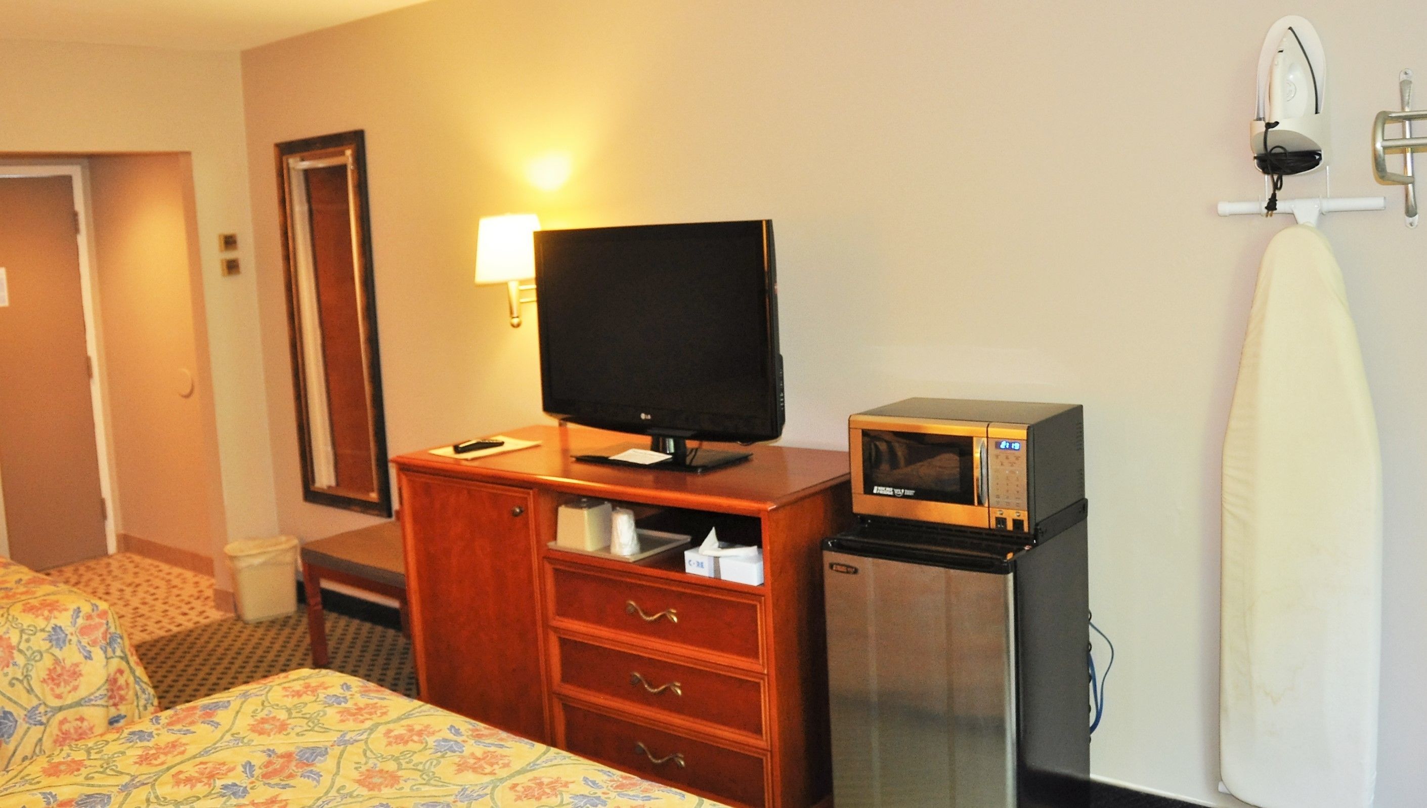Quarto de hotel com cômoda de madeira, televisão, micro-ondas e cama com colcha florida.
