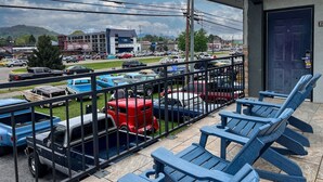 Varanda com cadeiras azuis, vista para um estacionamento com diversos veículos, e um prédio com uma porta escura ao fundo.
