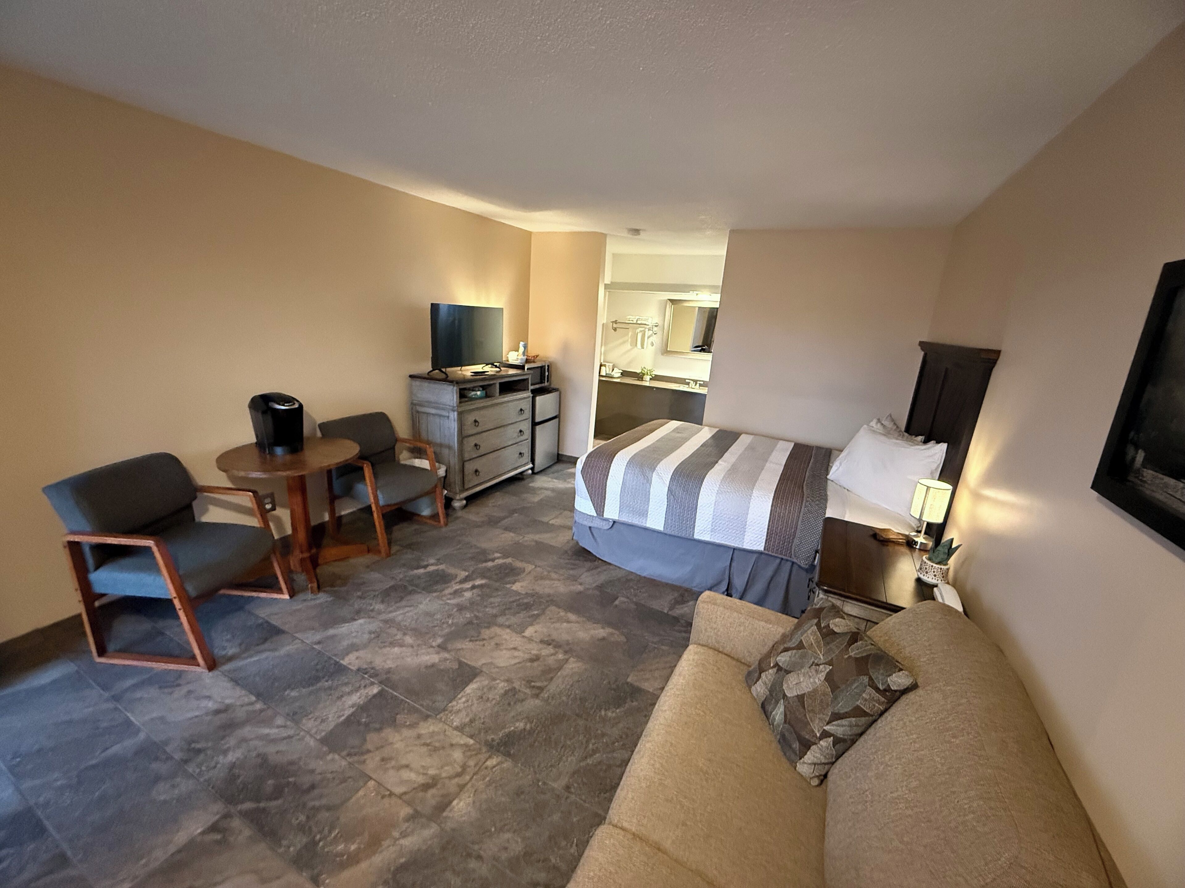 Quarto de hotel com uma cama, um sofá, uma escrivaninha com cadeira, uma televisão, um armário e um banheiro visível por uma porta aberta.