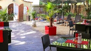 Uma área de pátio ao ar livre com mesas e cadeiras, algumas com detalhes vermelhos, e plantas em vasos.