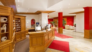 Um hall amplo com uma mesa de recepção de madeira, tapete vermelho e área de estar com uma cadeira branca.