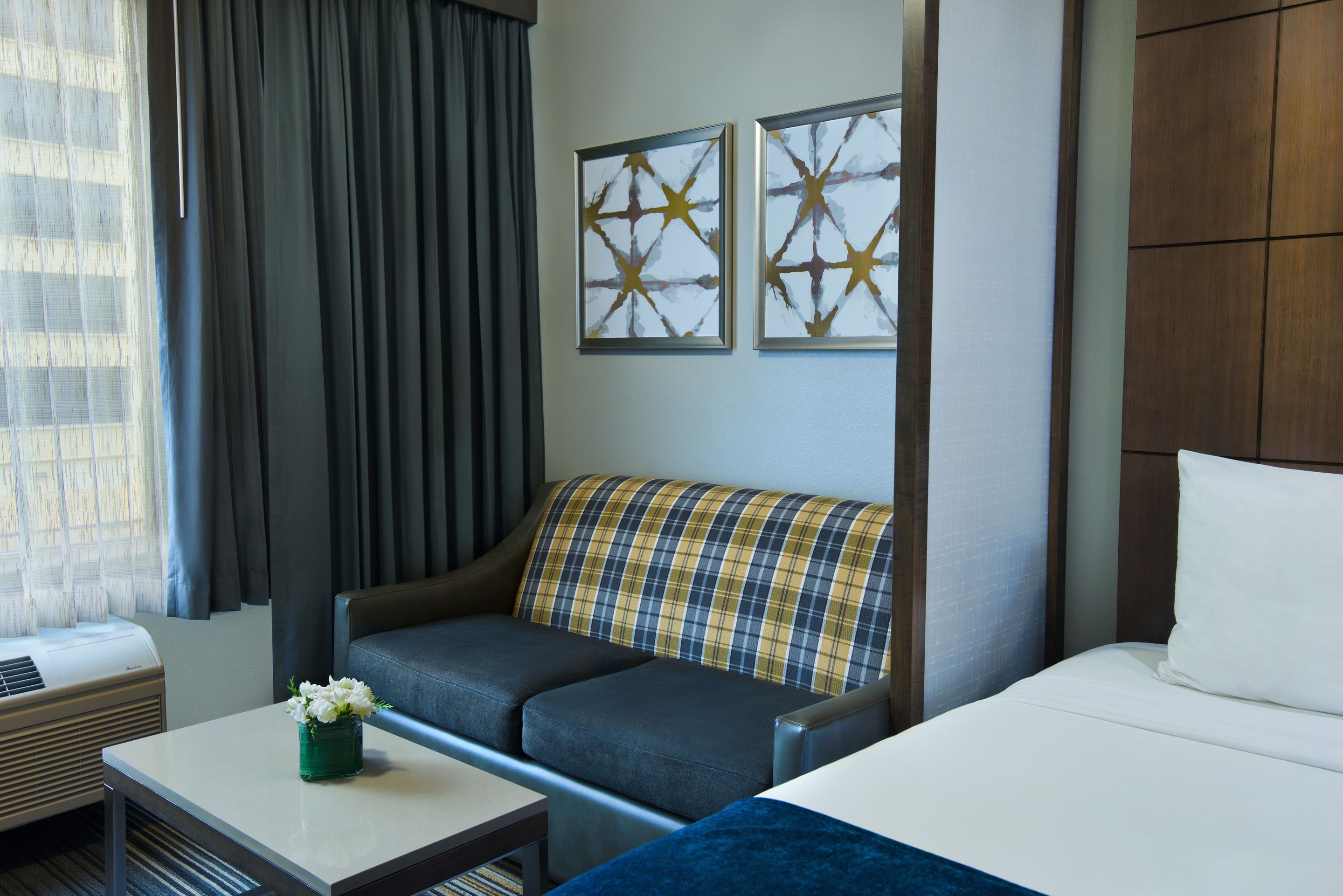 Quarto de hotel com cama, sofá, pequena mesa com um vaso de flores e duas obras de arte na parede.