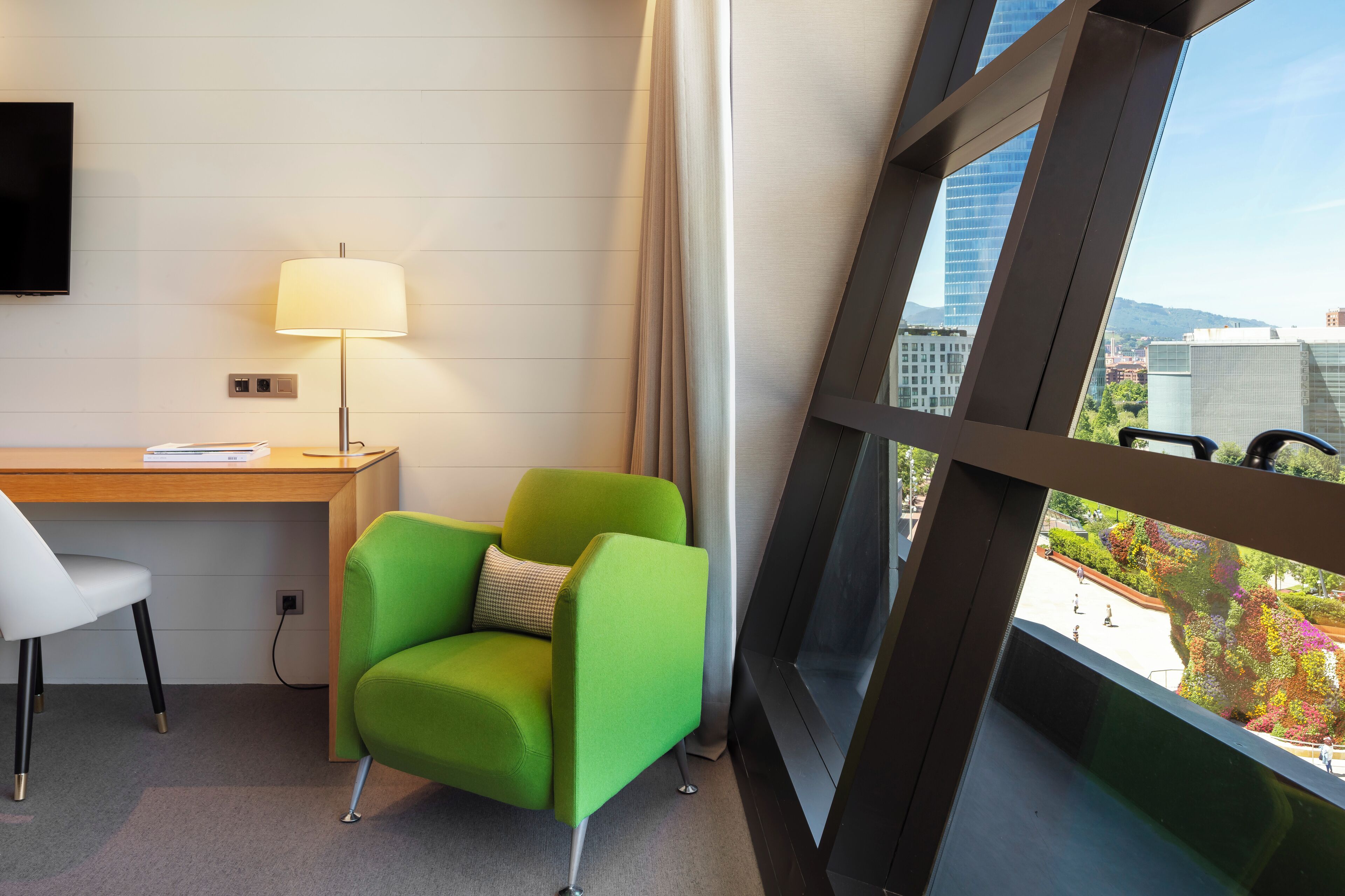 Quarto de hotel com escrivaninha, poltrona verde, luminária, televisão e janela com vista para a cidade.