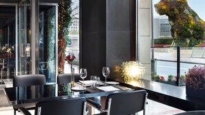 Uma sala de jantar moderna com mesa de vidro, cadeiras pretas e vista para um edifício coberto de flores.