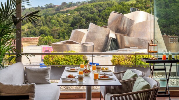 Uma sala de jantar com mesa posta para o café da manhã, vista para o teatro de ópera e varanda com plantas.