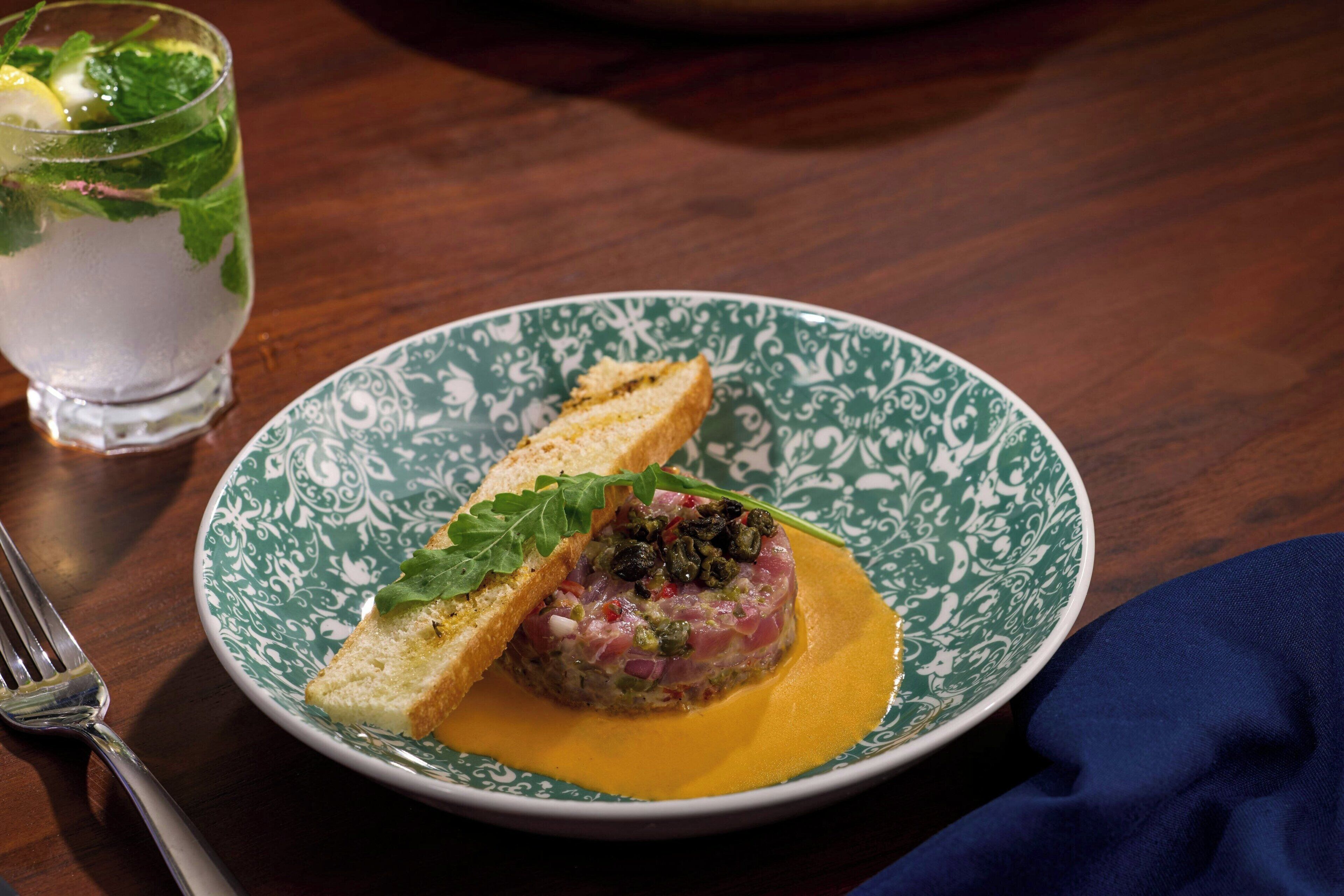 Um prato de steak tartare com torradas e molho, um copo de água com hortelã e um garfo sobre uma mesa de madeira.