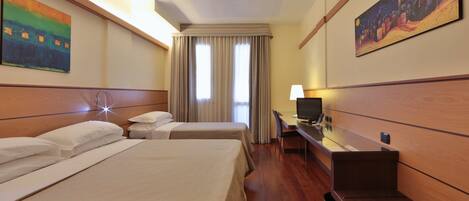 Quarto de hotel com duas camas, uma escrivaninha com computador, televisão e obras de arte nas paredes.