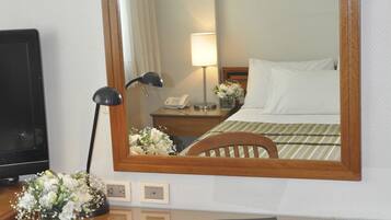 Quarto de hotel com escrivaninha de madeira, uma cadeira, cama com roupa branca, mesa de cabeceira com abajur e um espelho refletindo o quarto.