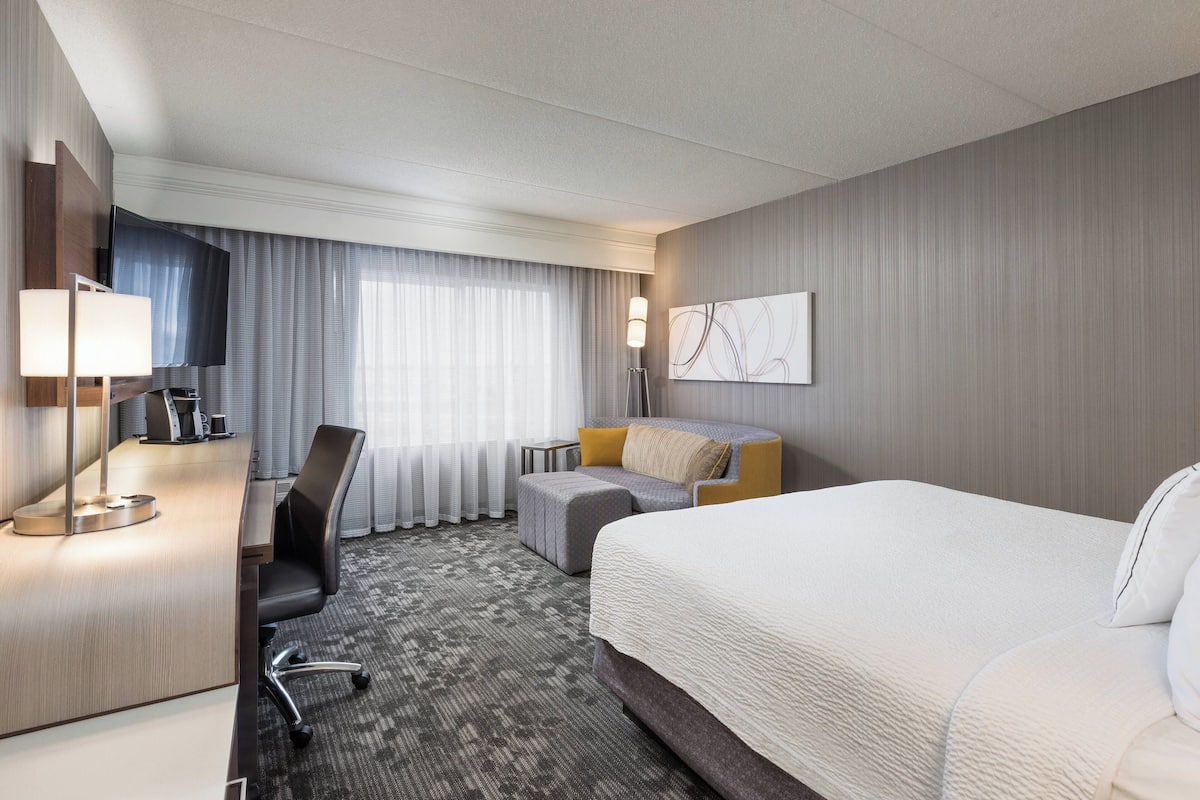 Quarto de hotel com uma cama grande, uma escrivaninha com abajur, um sofá e uma televisão.