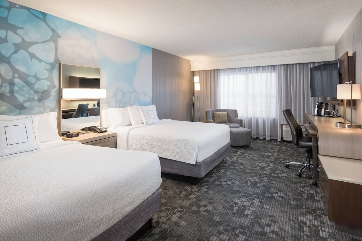 Quarto de hotel com duas camas, uma escrivaninha, uma cadeira, uma televisão e uma janela grande com cortinas.