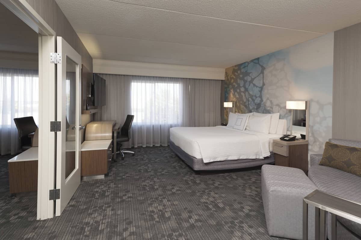 Quarto de hotel com cama, escrivaninha, cadeira, televisão e uma janela grande com cortinas.