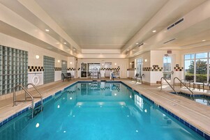 Uma piscina interior ampla com água azul cristalina, rodeada por um deck ladrilhado e com grades de metal.