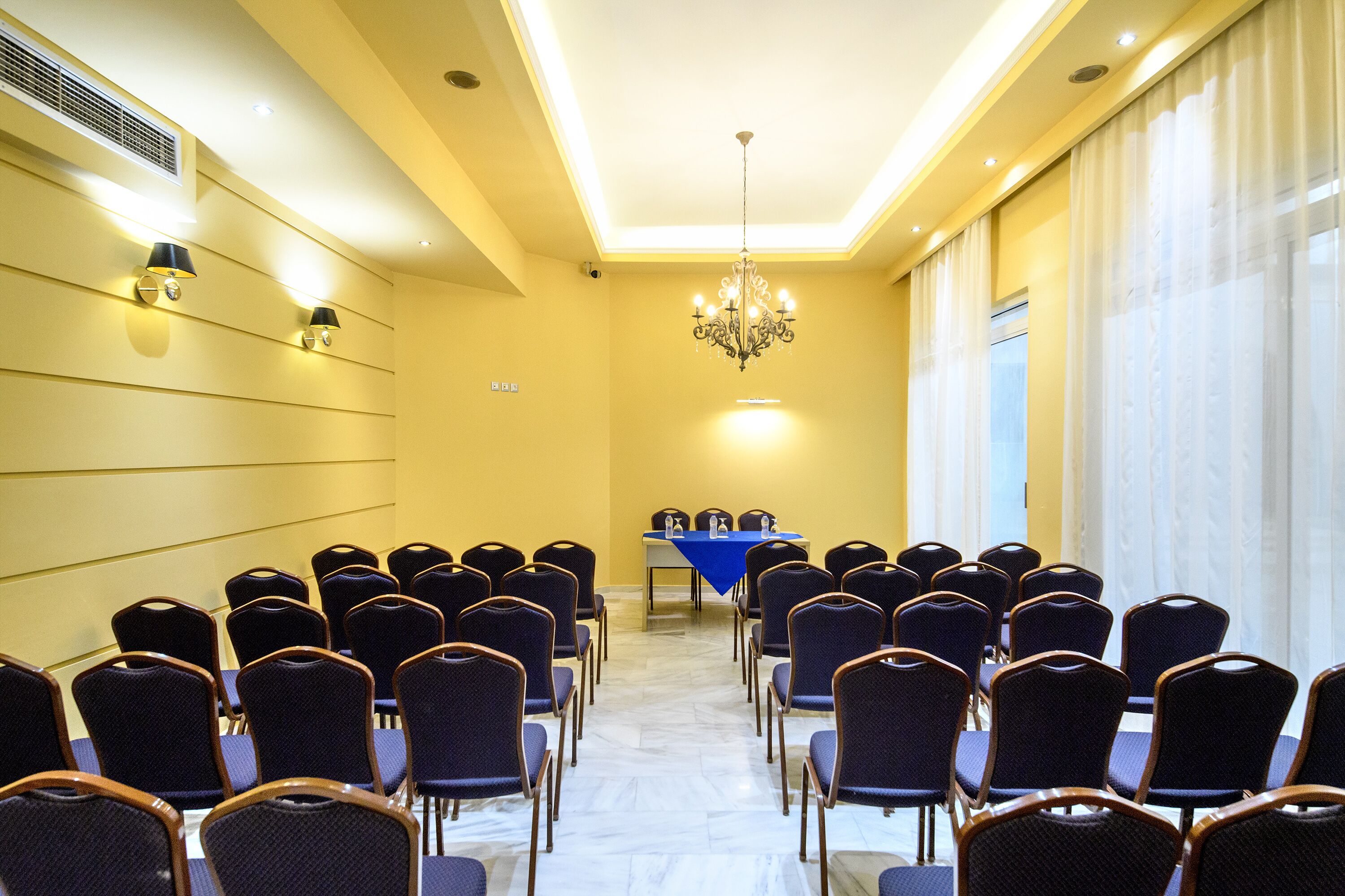 Sala de conferência com fileiras de cadeiras voltadas para uma mesa com toalha azul, um candelabro e luminárias fixadas na parede.