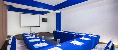 Sala de conferências com uma mesa comprida, cadeiras brancas e uma tela de projeção.