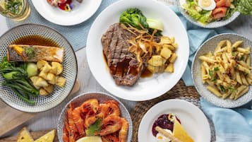Diversos pratos, incluindo bife, camarão e massa, dispostos sobre uma mesa com toalha azul.