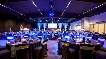 Uma sala de conferências ampla, com várias mesas redondas, cadeiras azuis e um palco com uma tela na frente.