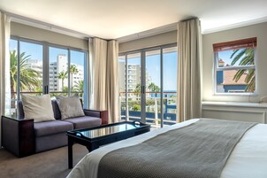 Quarto de hotel com uma cama grande, um sofá, uma mesa de centro de vidro e vista para palmeiras e edifícios através de grandes janelas.