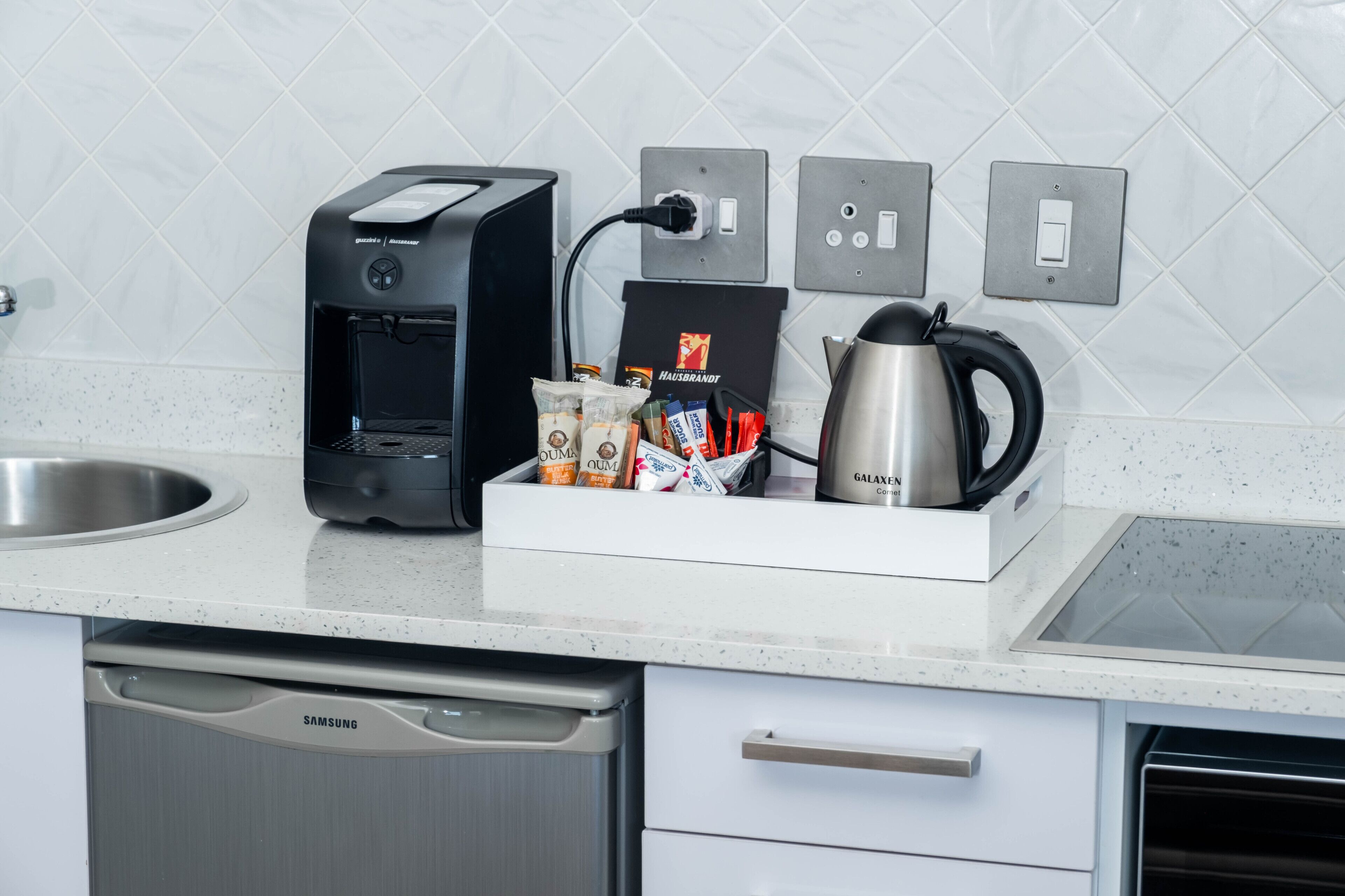 Uma cozinha com cafeteira, chaleira e suporte para cápsulas de café no balcão.