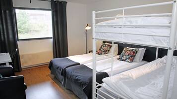 Quarto com duas beliches, uma escrivaninha e uma cadeira. Há uma janela com cortinas, um abajur e um radiador.
