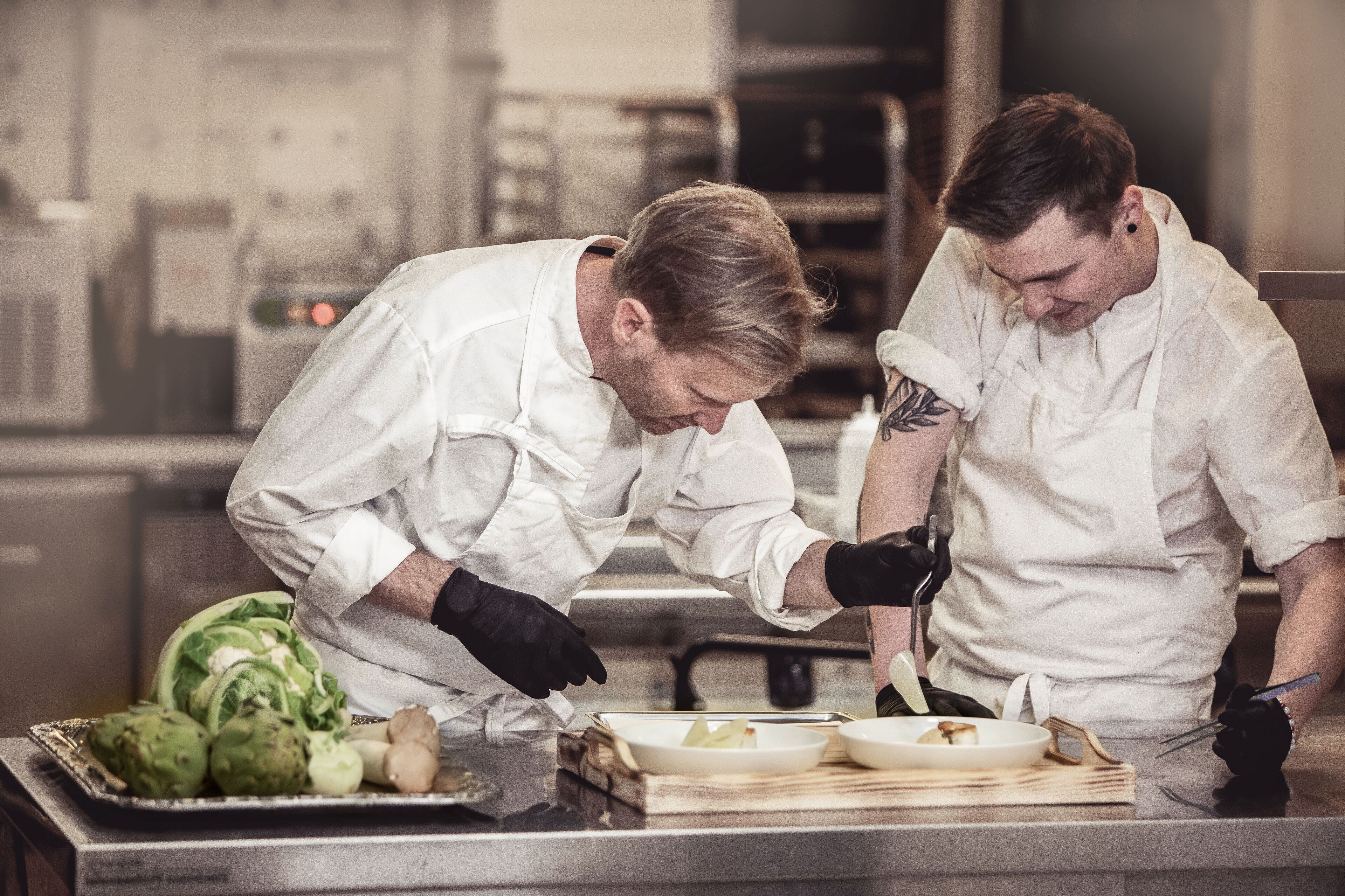 Dois chefs preparando alimentos em uma cozinha profissional com superfícies de aço inoxidável e equipamentos modernos.