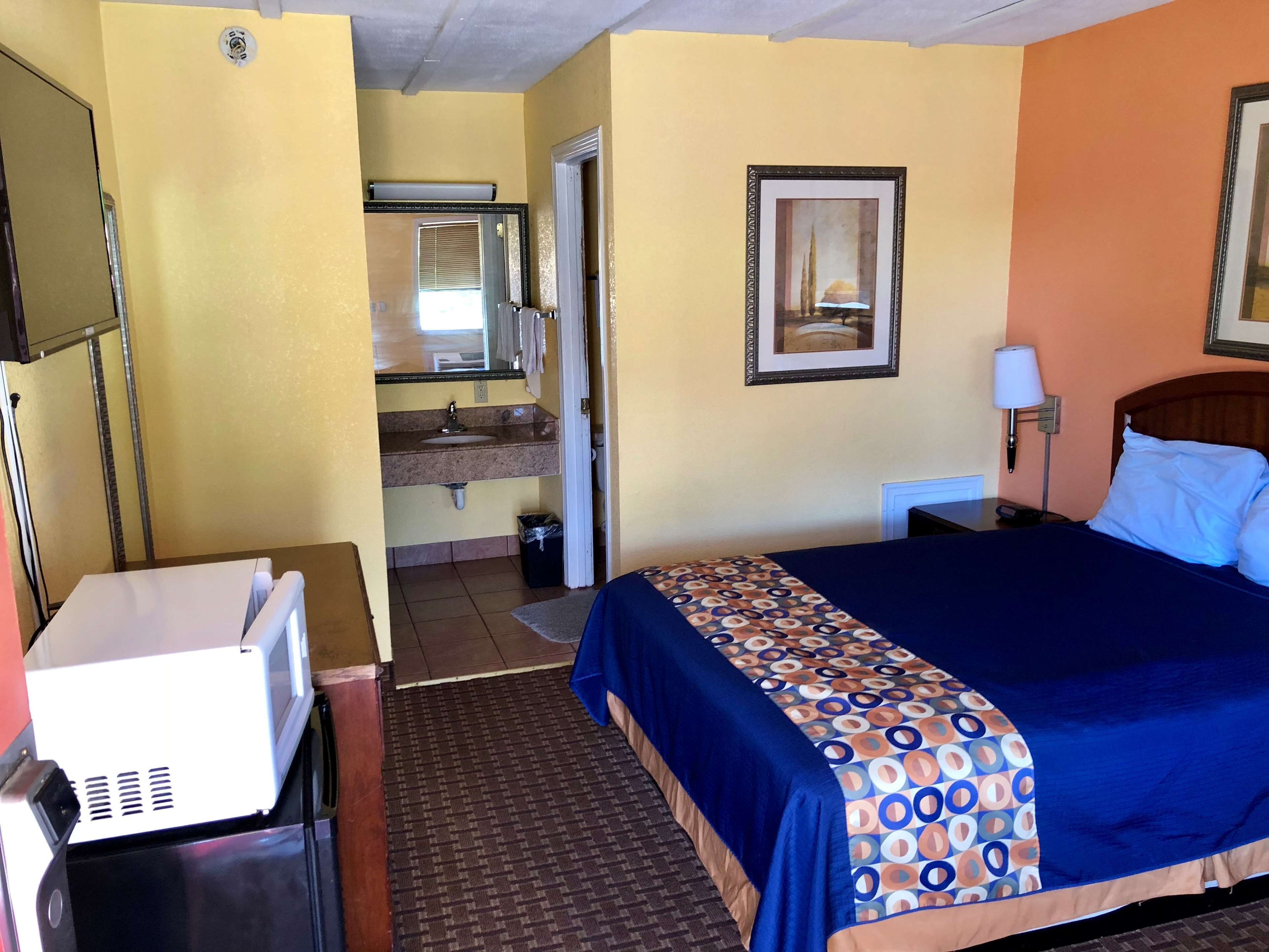 Quarto de hotel com cama, escrivaninha, cadeira, abajur, quadro na parede e banheiro visível por uma porta aberta.