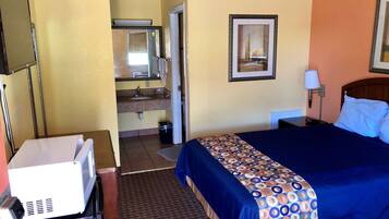 Quarto de hotel com cama, escrivaninha, cadeira, abajur, quadro na parede e banheiro visível por uma porta aberta.