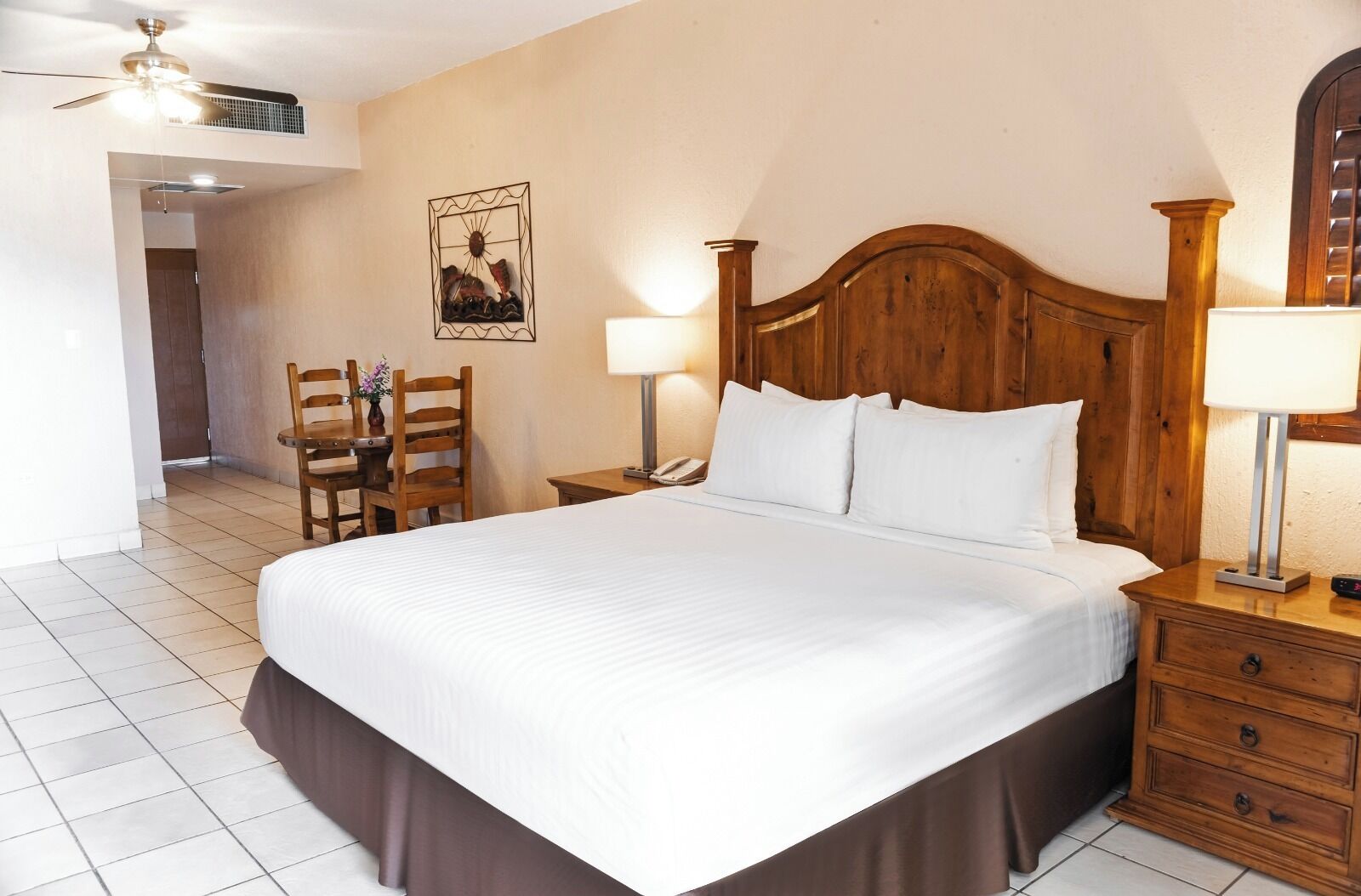Quarto de hotel com uma cama grande, cabeceira de madeira, duas mesas de cabeceira com luminárias, uma cadeira de madeira e uma obra de arte na parede.