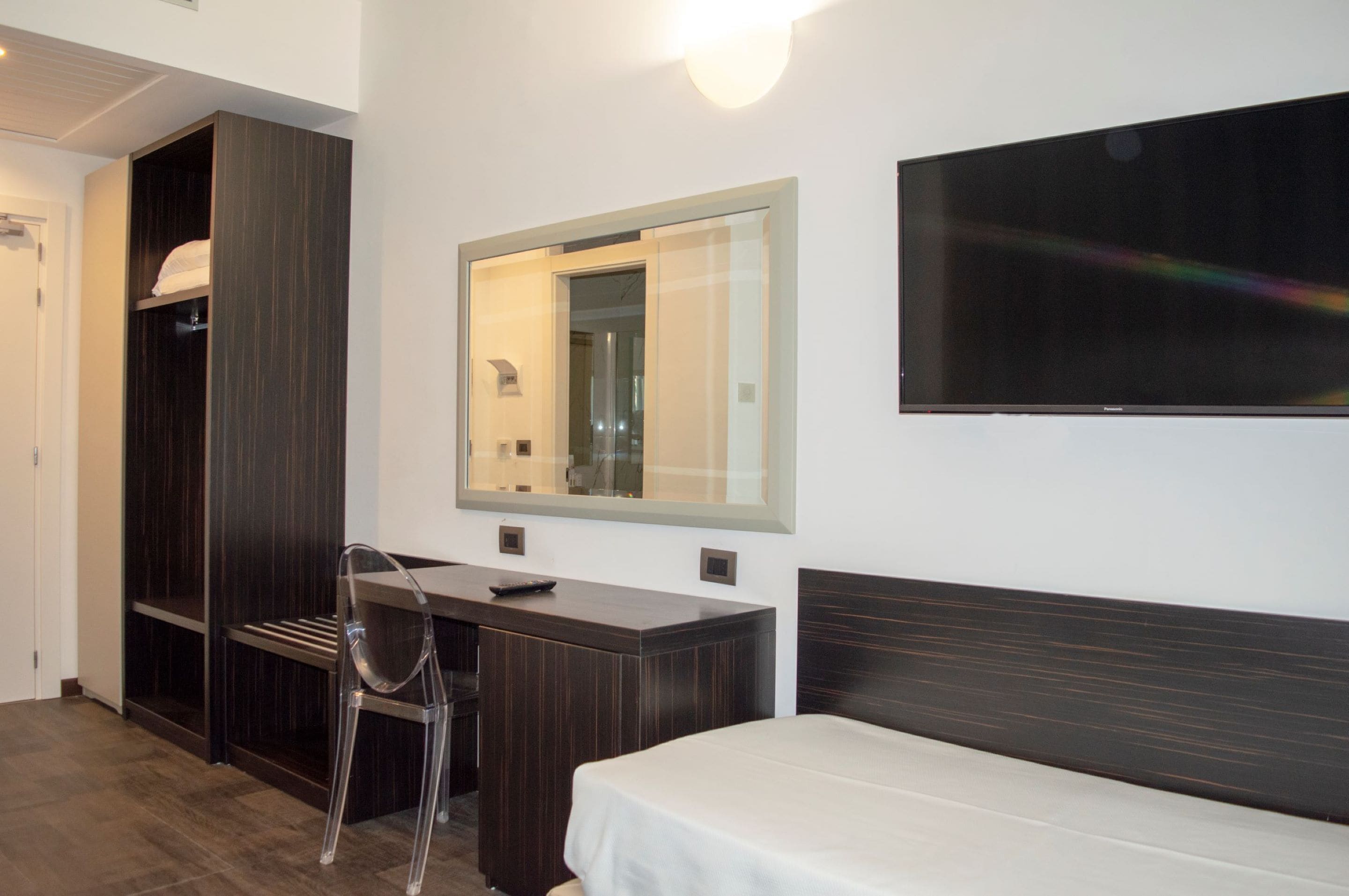 Quarto de hotel com cama, escrivaninha, cadeira, guarda-roupa e televisão na parede.