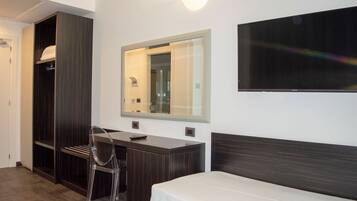 Quarto de hotel com cama, escrivaninha, cadeira, guarda-roupa e televisão na parede.