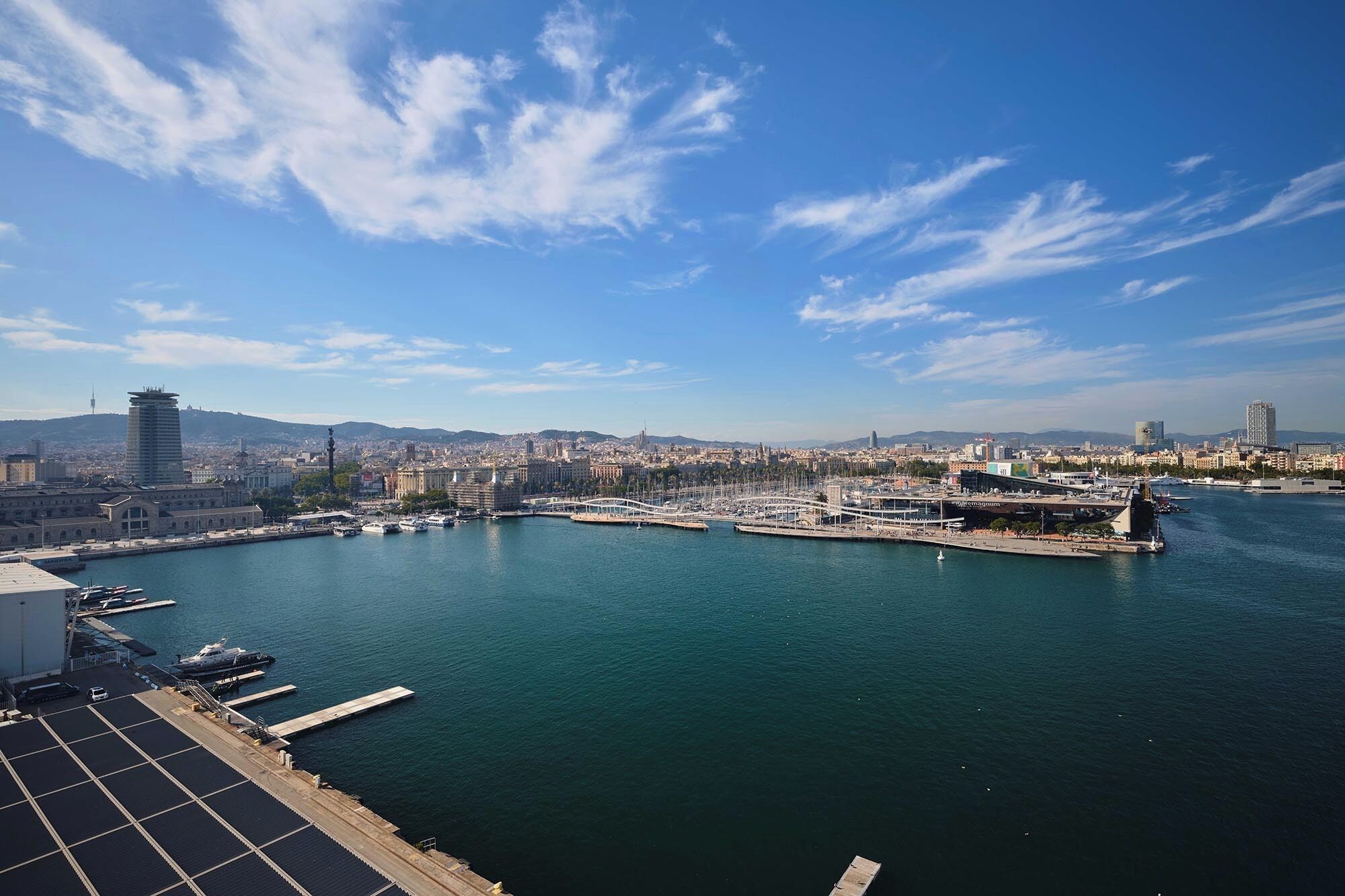 Uma marina com barcos atracados, um edifício de grandes dimensões com design singular e um horizonte urbano ao fundo.