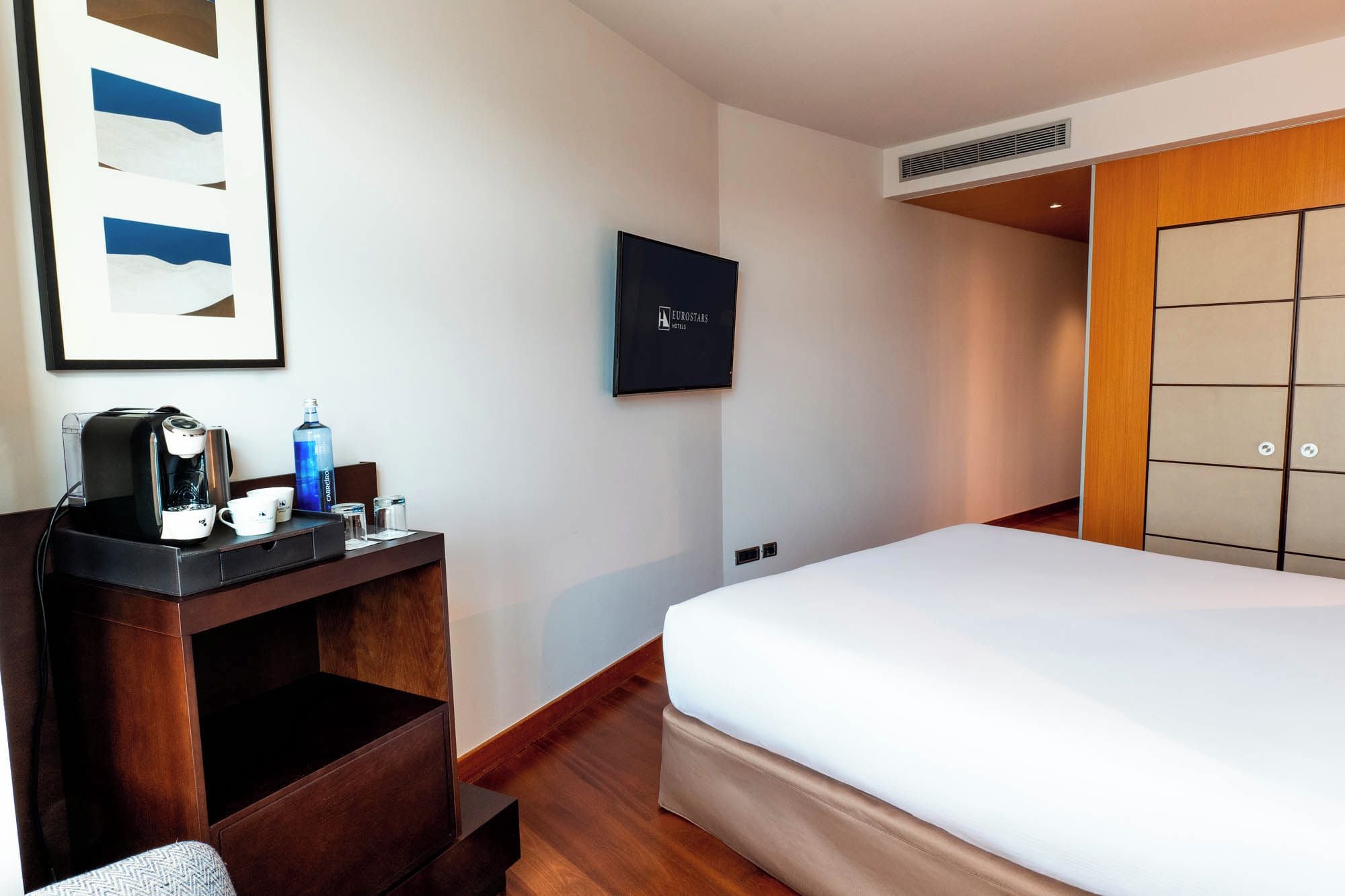 Quarto de hotel com cama, escrivaninha com cafeteira, televisão e minibar.