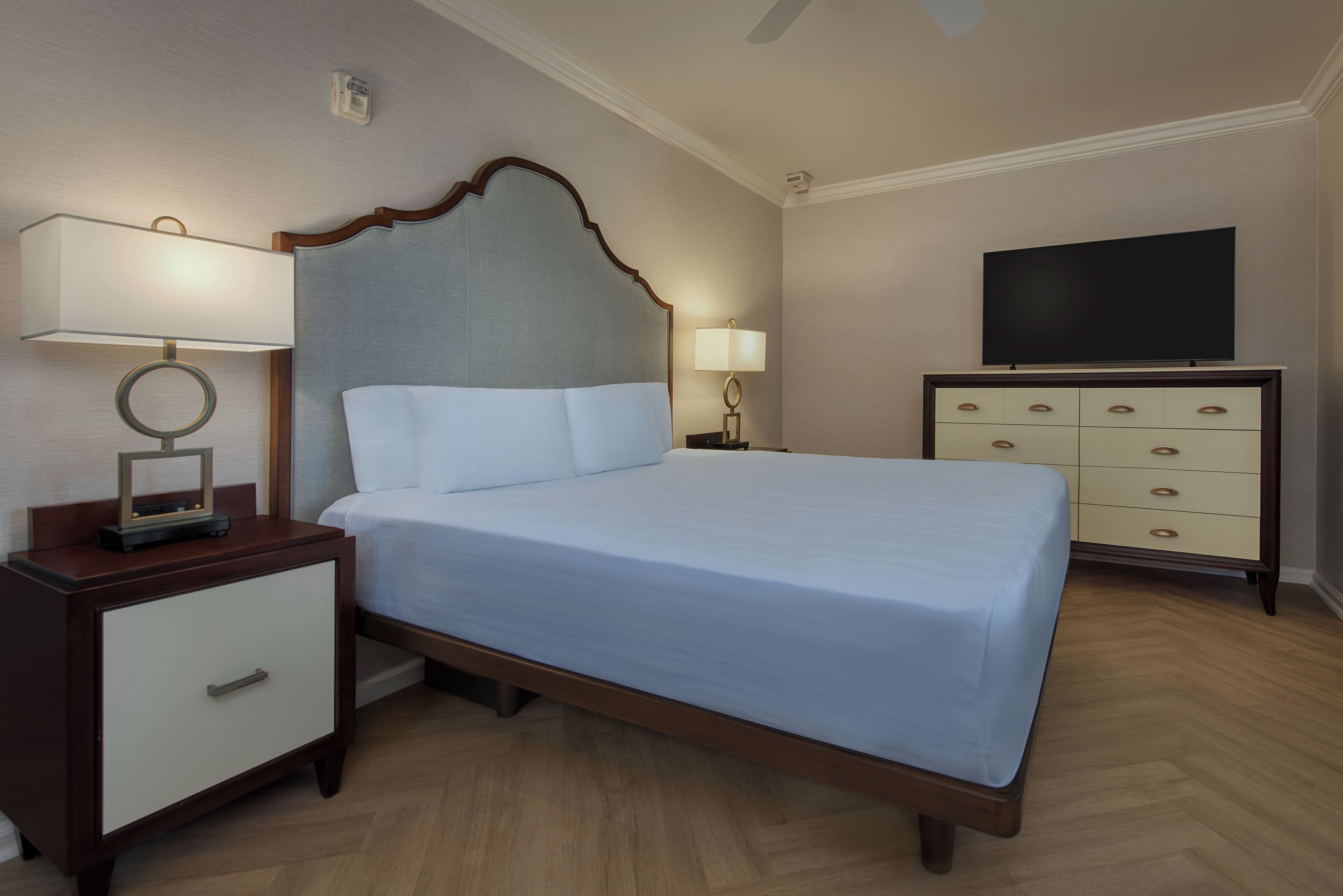 Quarto de hotel com cama, mesas de cabeceira com luminárias, televisão e cômoda.