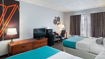 Quarto de hotel com duas camas, uma mesa com computador, televisão e parede com decoração geométrica.