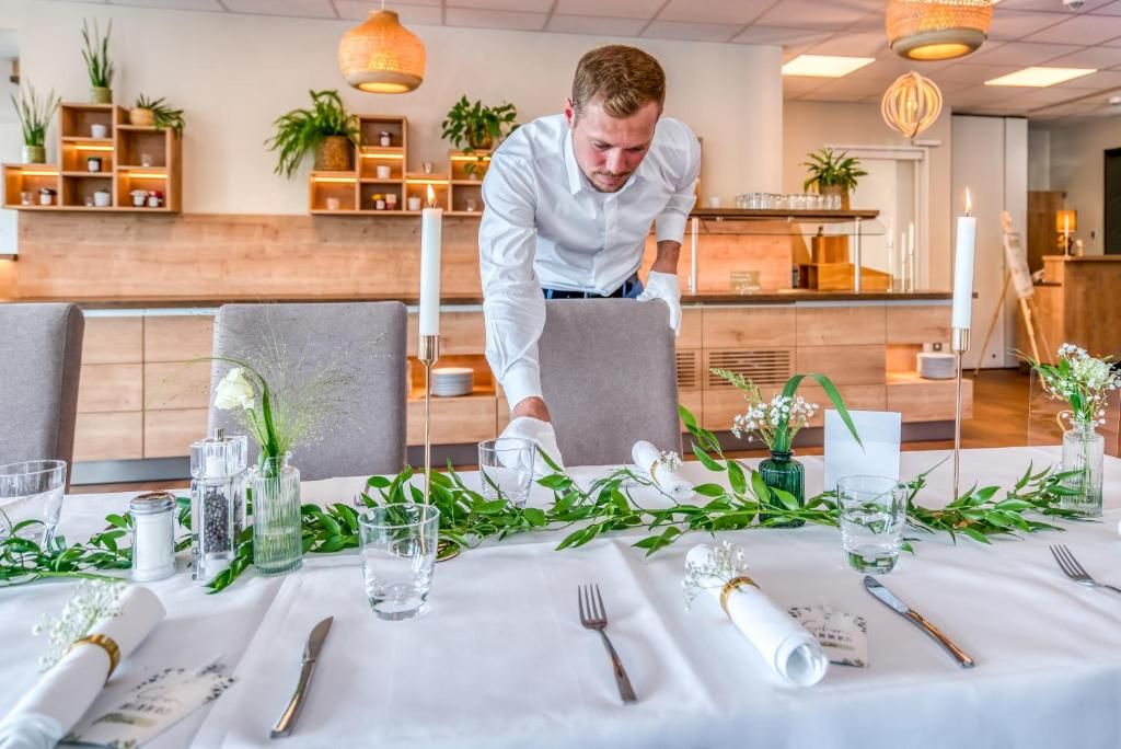 Mesa de jantar bem posta, com toalha branca, talheres organizados e uma pessoa cuidando da mesa.