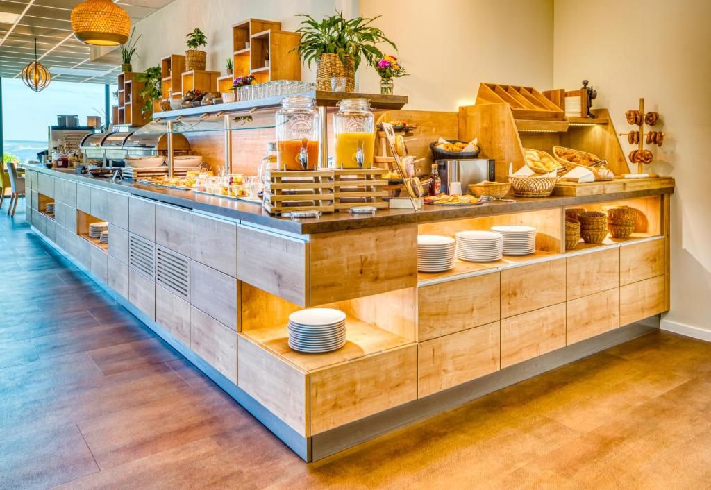 Disposição de buffet com diversos alimentos, um espaço para café e um balcão com pilhas de pratos.