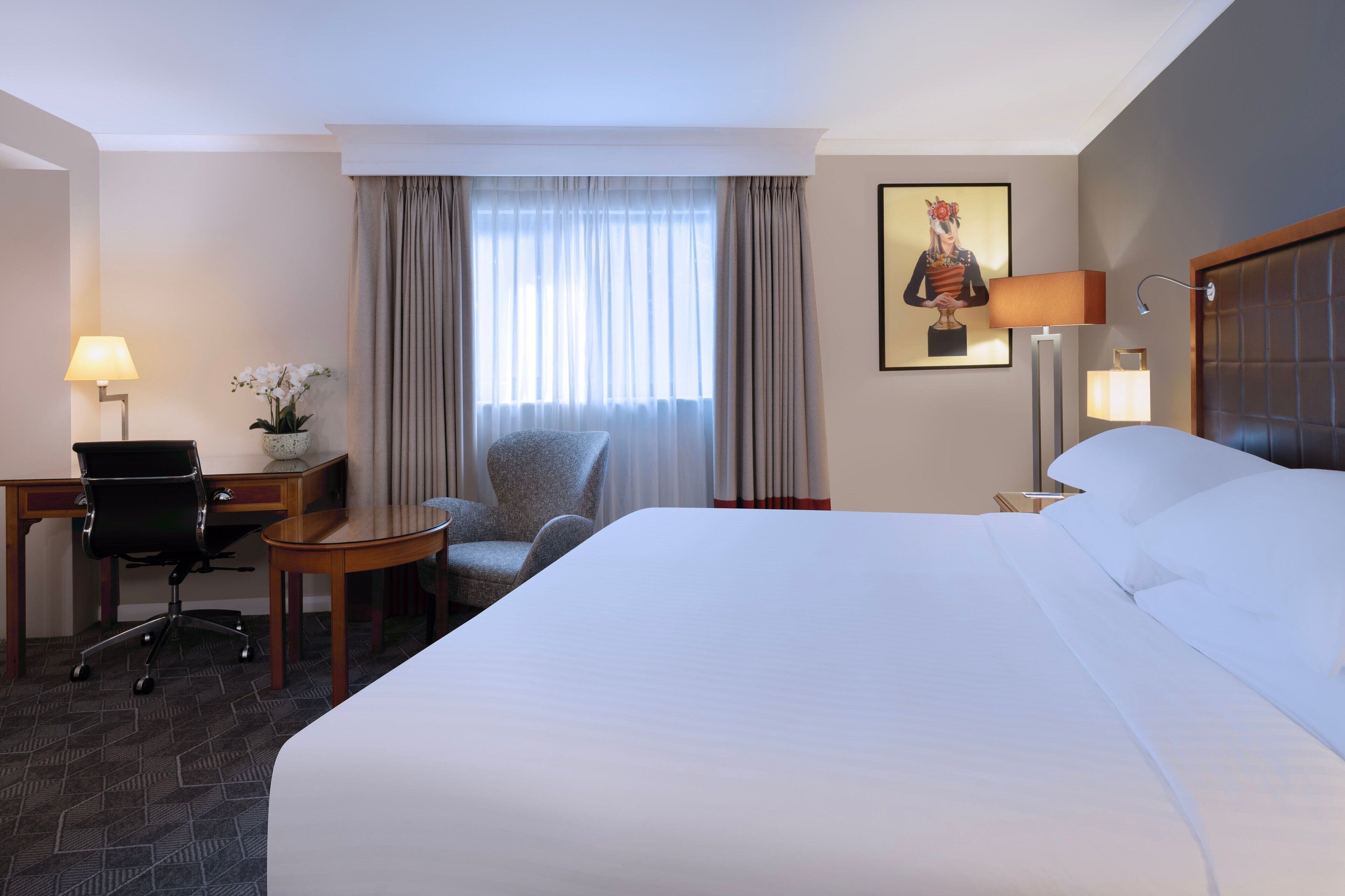 Quarto de hotel com uma cama grande, uma mesa com cadeira, uma luminária e um quadro na parede.