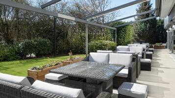 Um pátio externo com móveis de vime, uma mesa de madeira e uma estrutura de pérgola.