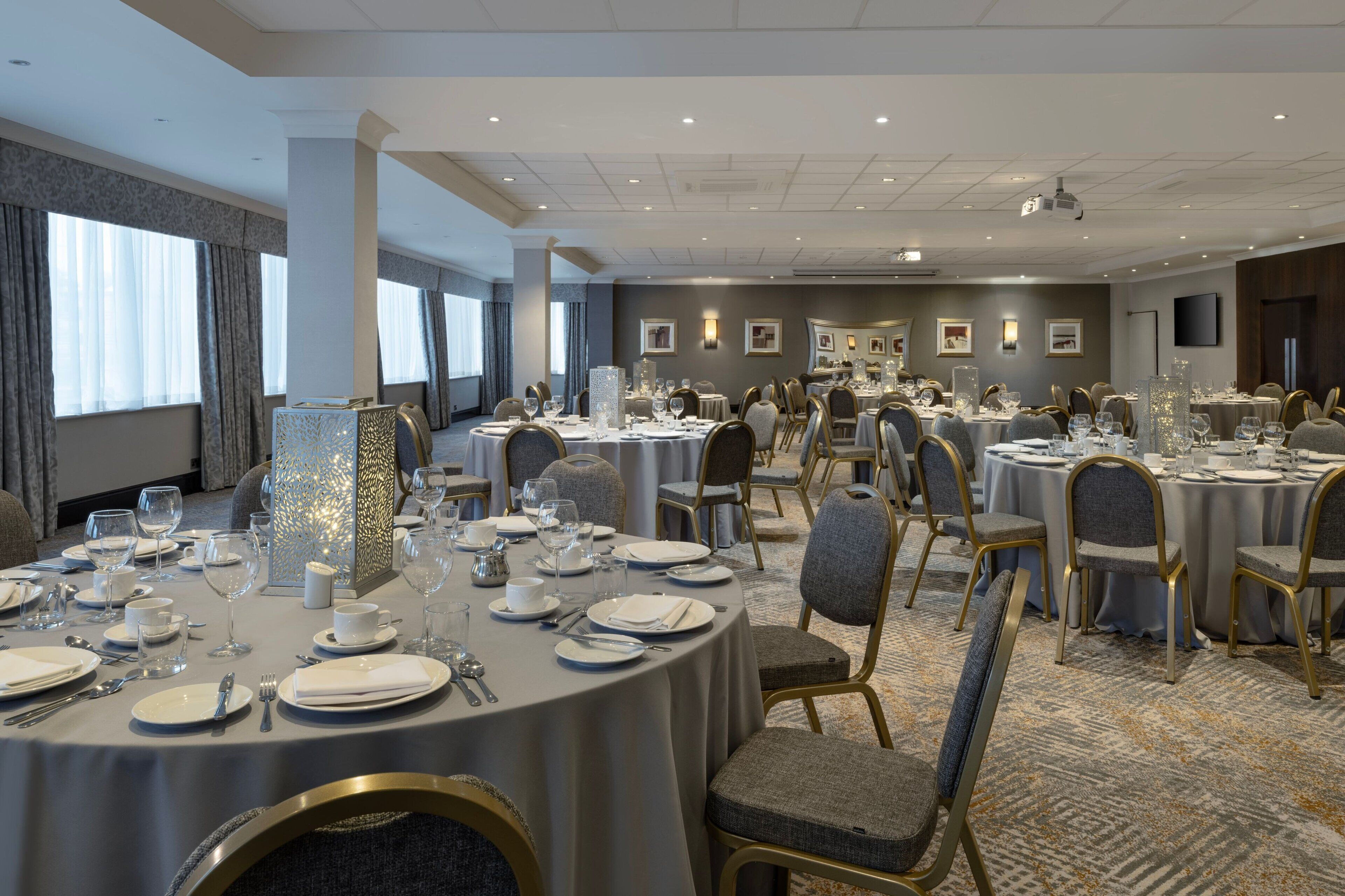 Uma sala de banquetes com mesas redondas preparadas para refeições, equipada com cadeiras, utensílios de mesa e elementos decorativos.