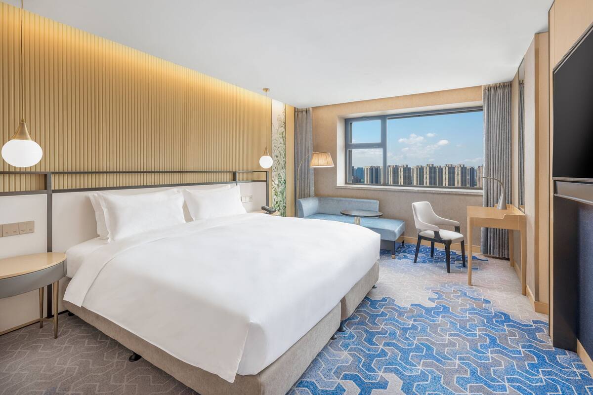 Quarto de hotel com uma cama grande, uma escrivaninha, uma cadeira, televisão e janela com vista para a cidade.