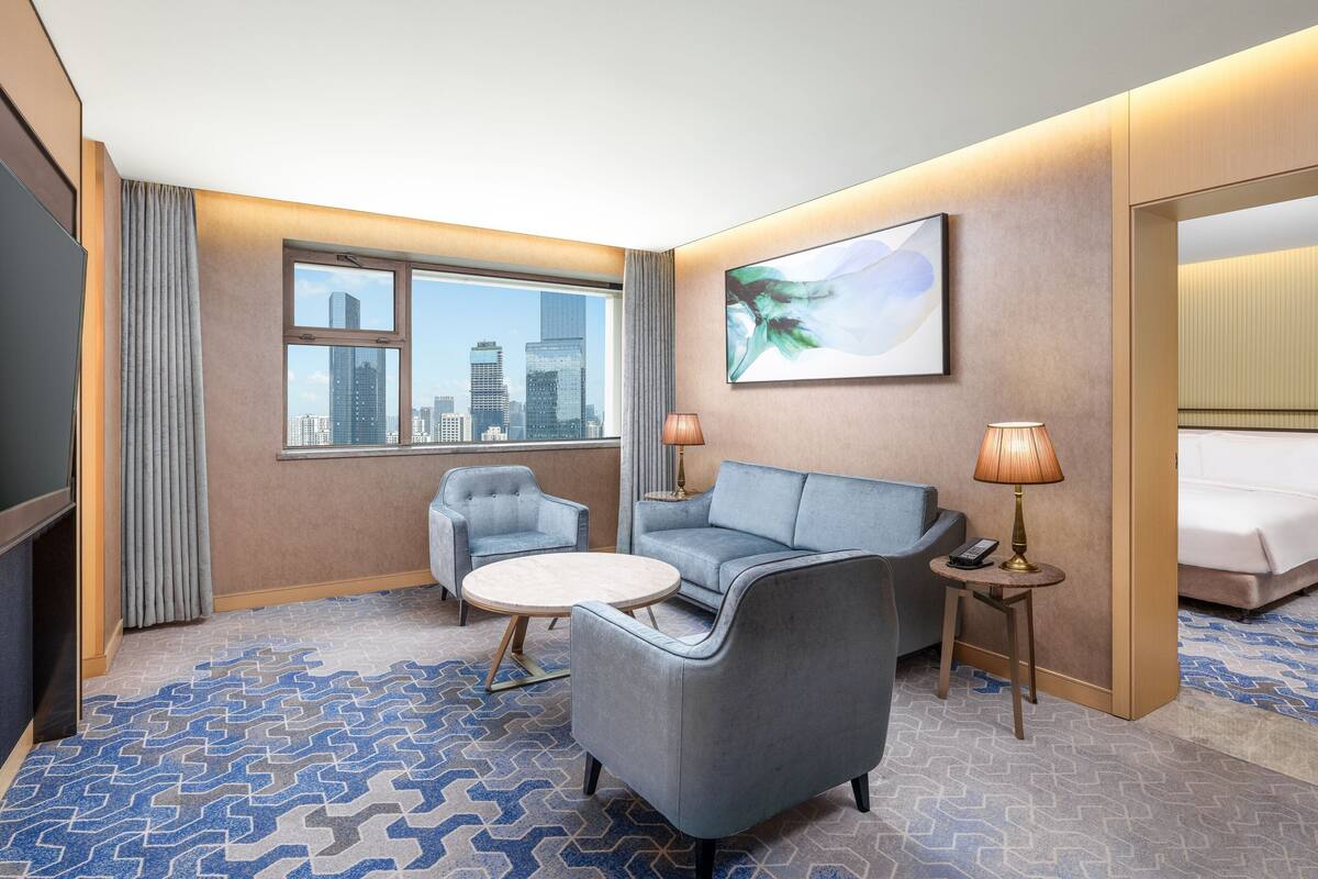 Quarto de hotel com uma janela grande, um sofá, uma poltrona, uma mesa pequena e um abajur.