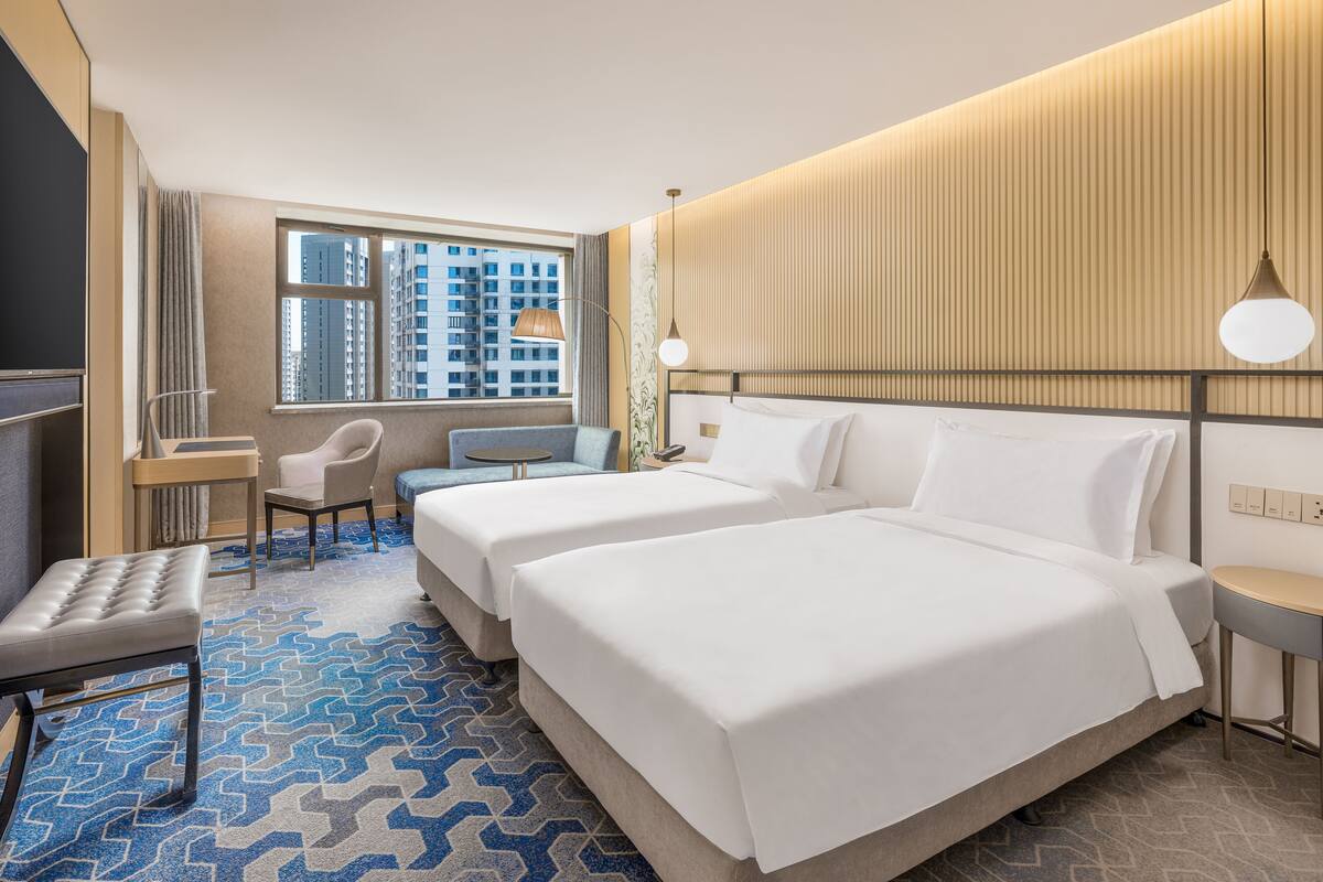 Quarto de hotel com duas camas, uma escrivaninha, uma cadeira e vista para edifícios altos.