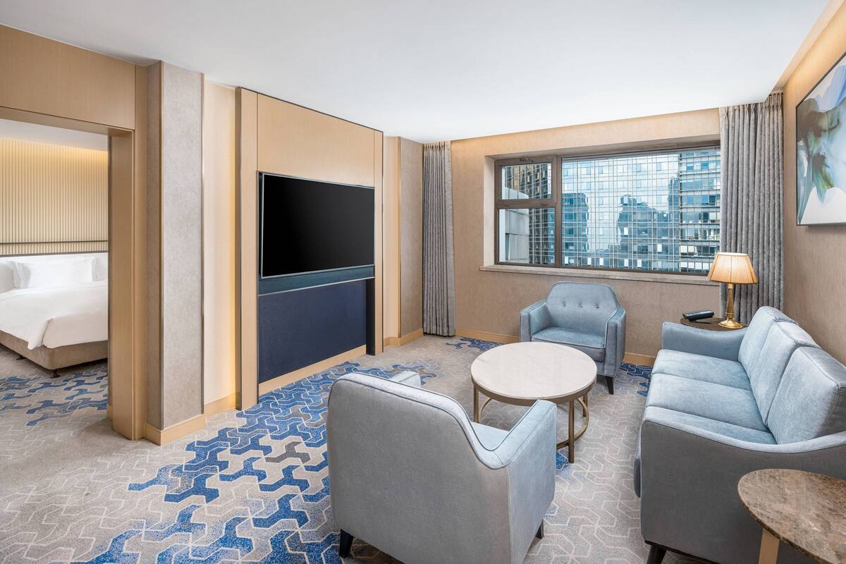 Quarto de hotel com cama, televisão de tela plana, sofá, poltrona, mesa pequena e janela com vista para a cidade.