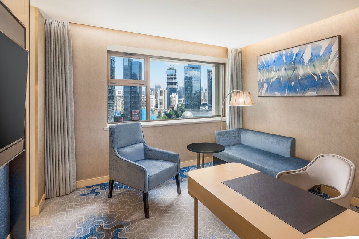 Quarto de hotel com vista para a cidade, um sofá, uma cadeira, uma mesinha e um abajur.