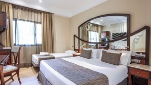 Quarto de hotel com duas camas, uma escrivaninha, uma cadeira, um espelho grande e uma janela com cortinas.