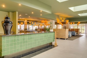 Um saguão de hotel com uma mesa de recepção, várias luminárias de vime suspensas e uma grande planta em vaso.