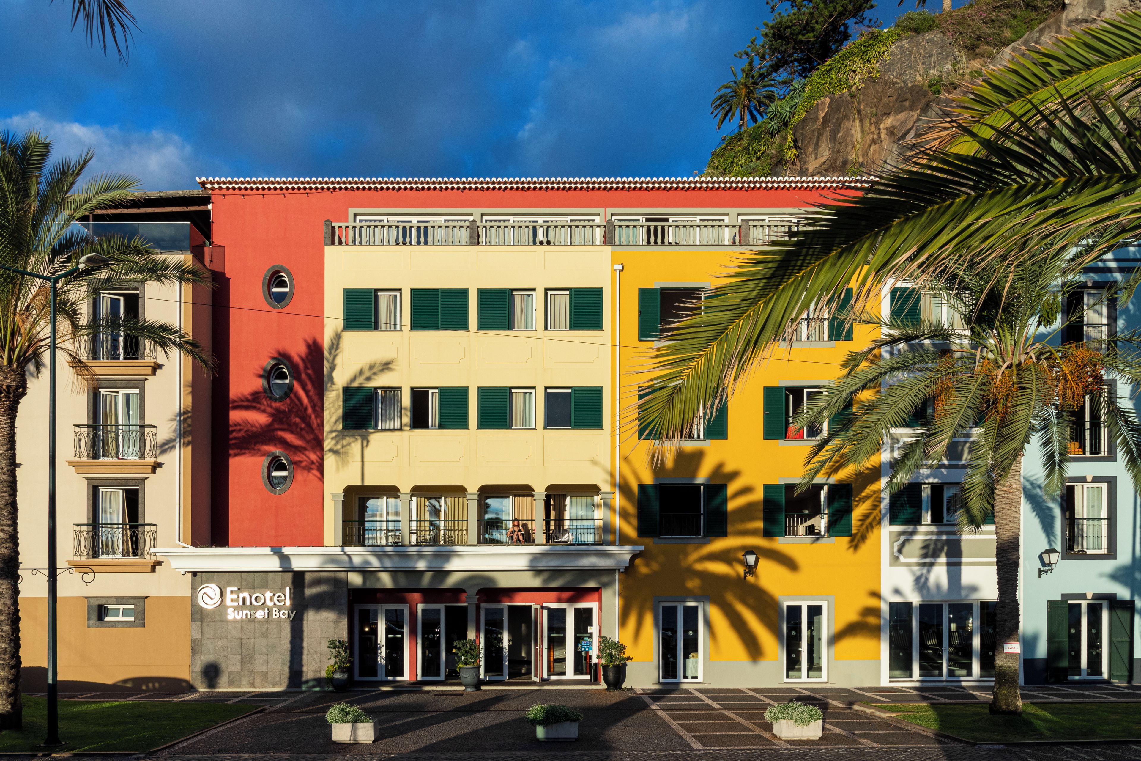 Um hotel com fachada em vermelho, amarelo e azul, várias sacadas e palmeiras.